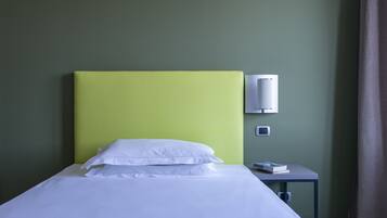 Quarto de hotel com cabeceira verde, roupa de cama branca, mesa de cabeceira com um livro e uma luminária na parede.