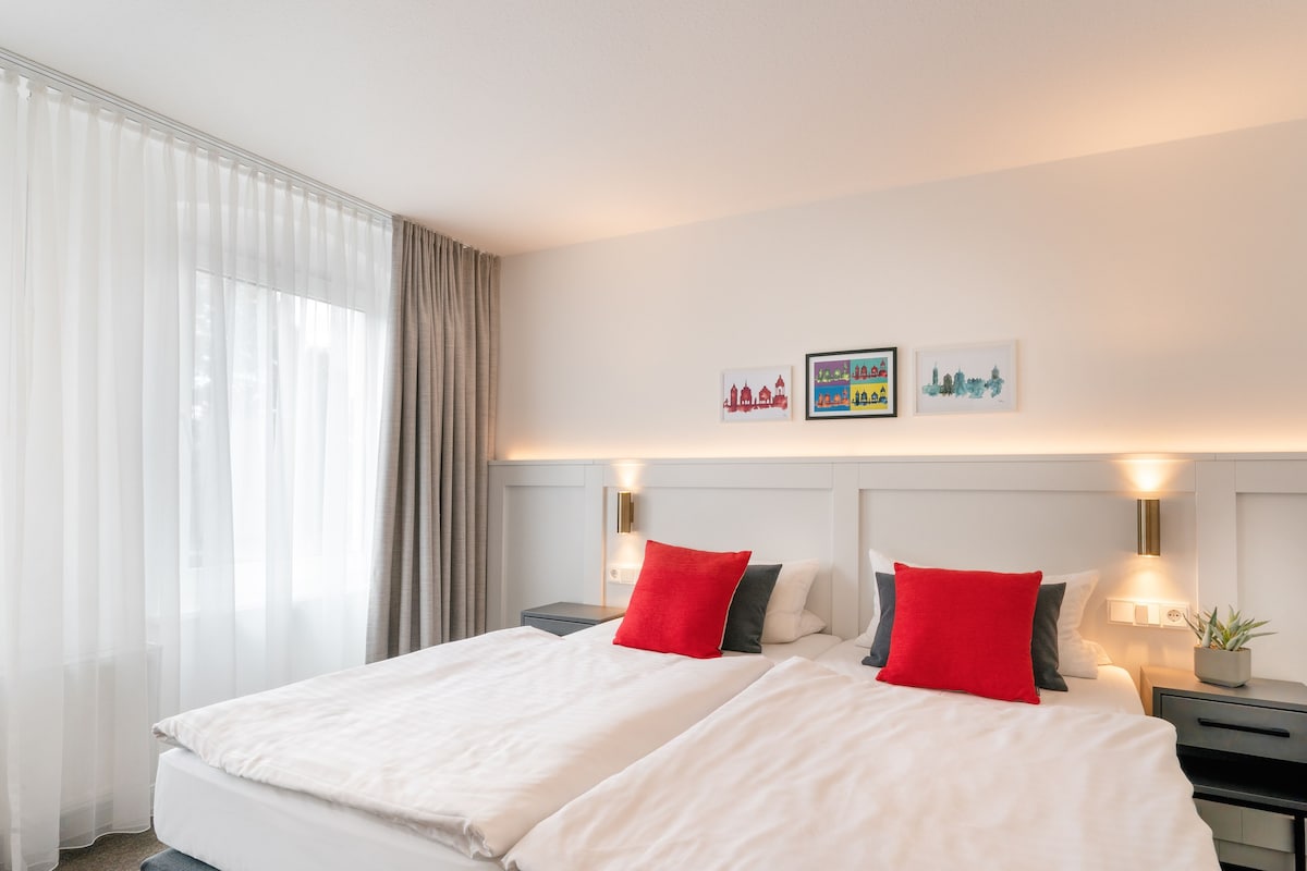 Quarto de hotel com uma cama grande, duas mesas de cabeceira e uma janela com cortinas leves.