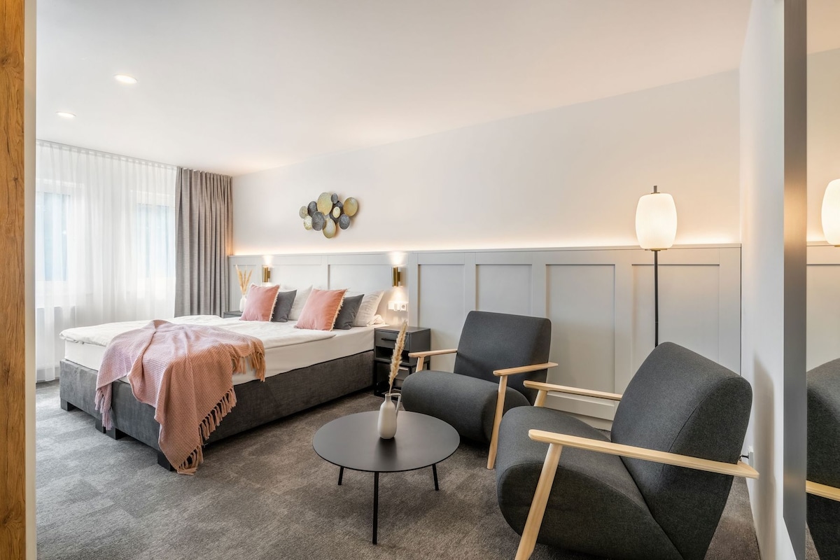 Quarto de hotel moderno com uma cama, dois poltronas, uma mesa de centro e uma luminária de parede.