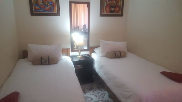 Quarto de hotel com duas camas de solteiro, uma mesa de cabeceira com luminária e obras de arte nas paredes.