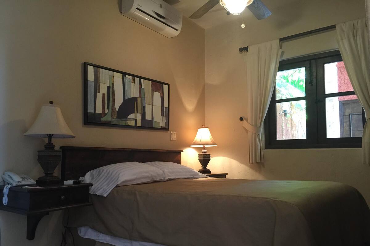 Um quarto com cama, mesa de cabeceira com abajur, pintura e janela com cortinas.