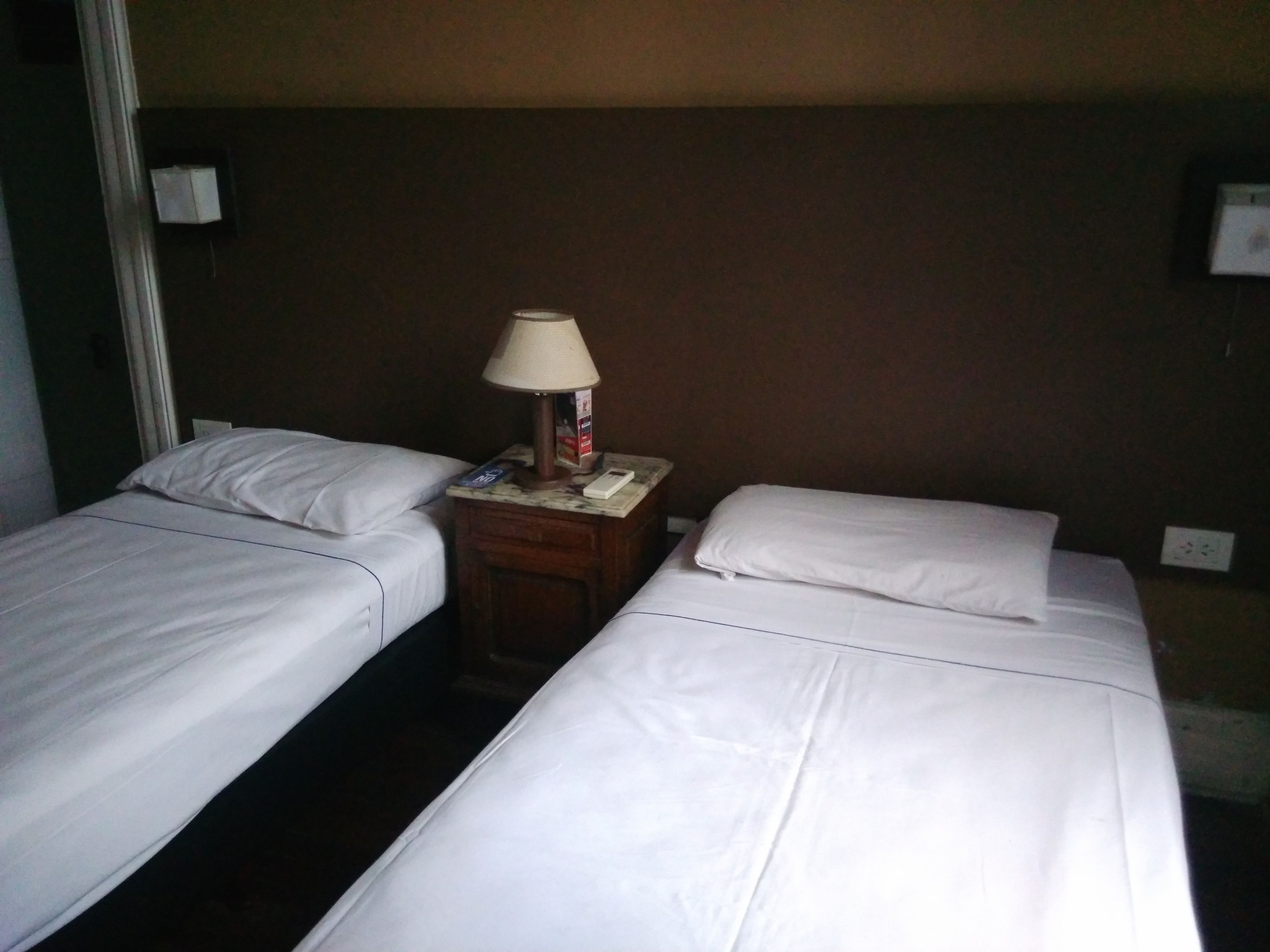 Quarto de hotel com duas camas de solteiro, uma mesa de cabeceira com abajur e uma pequena mesa.