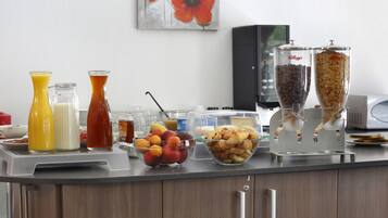 Um balcão de pequeno-almoço com diversos alimentos e bebidas, incluindo uma taça de fruta, um copo de sumo de laranja e um recipiente com cereais.