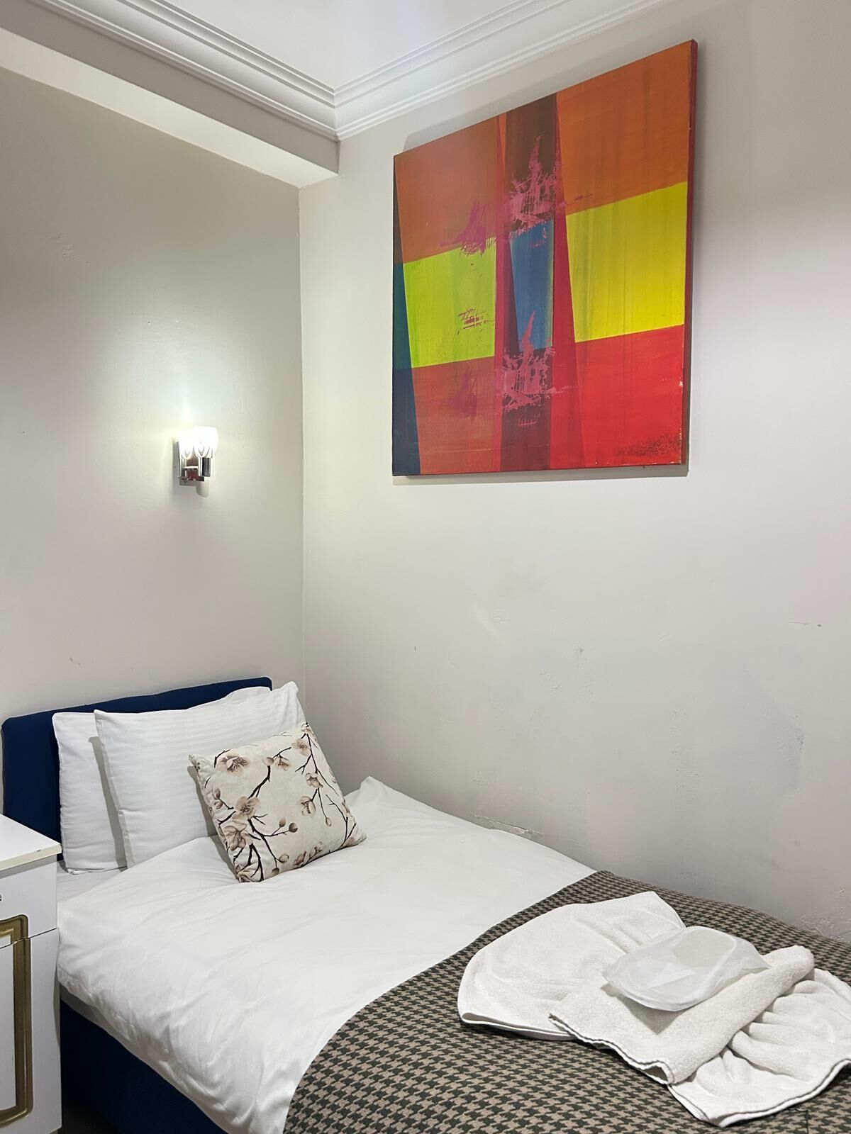 Quarto de hotel com cama bem arrumada, luminária fixada na parede e pintura abstrata colorida.