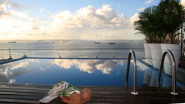 Piscina com vista clara do oceano e navios, deck de madeira e planta em vaso.
