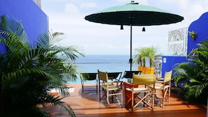 Um deck de madeira com uma mesa, cadeiras, um guarda-sol verde e vista para o mar.