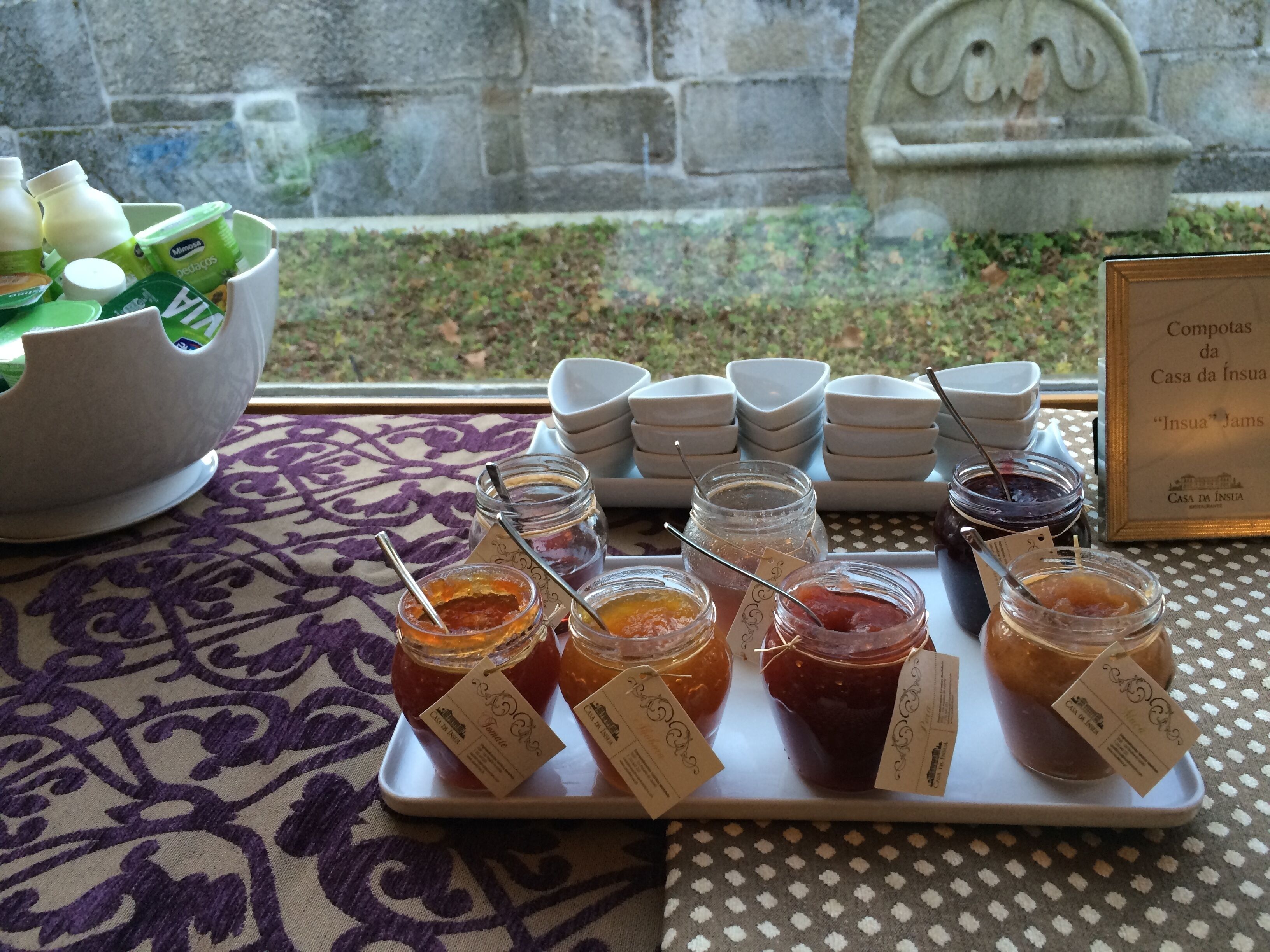 Uma mesa com potes de geleia e uma tigela de iogurte, um letreiro com os doces 'Casa da Insua', e um muro de pedra ao fundo.