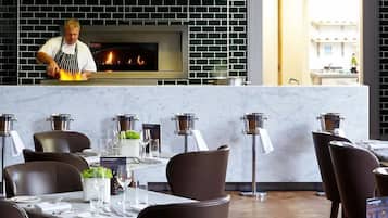 Um restaurante com parede de tijolos e forno a lenha. Mesas com toalhas brancas e cadeiras marrons.