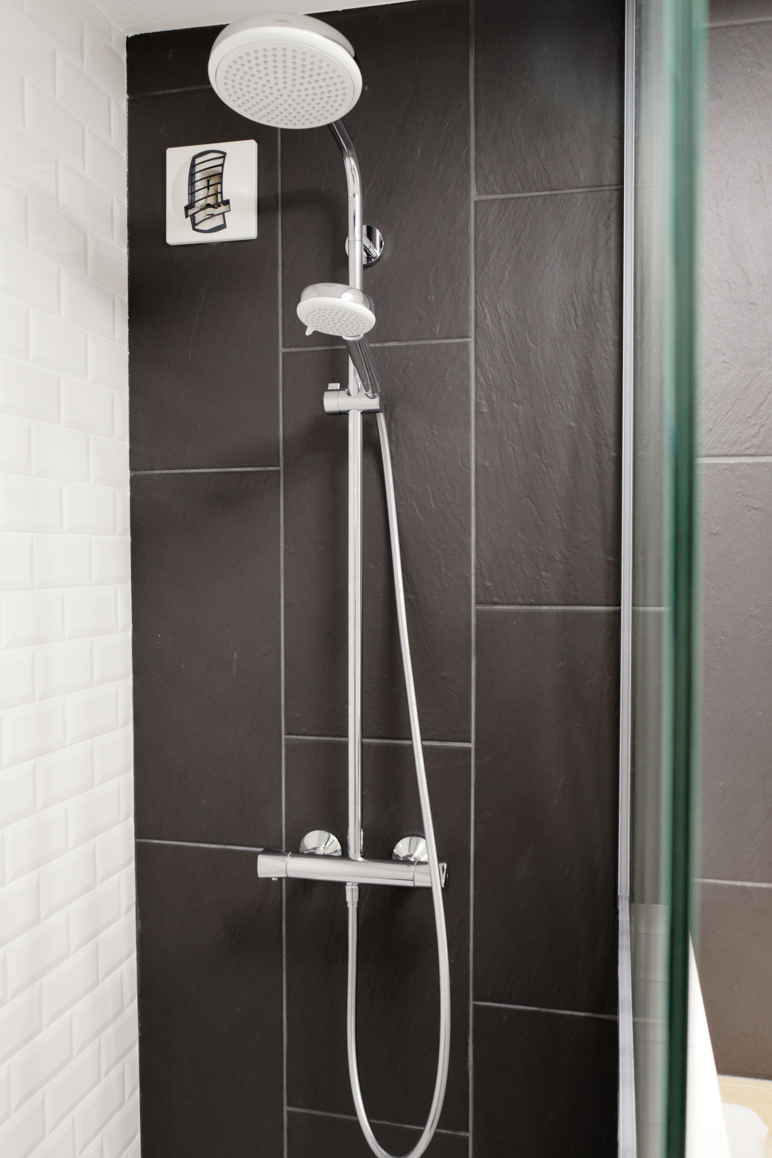 Chuveiro com chuveiro manual e fixo, instalado numa parede com azulejos escuros.