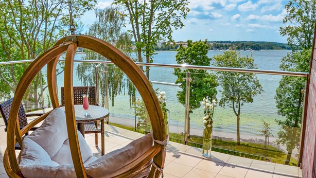Varanda com cadeira e mesa de vime, com vista para um lago, árvores e uma linha de costa distante.