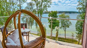 Varanda com cadeira e mesa de vime, com vista para um lago, árvores e uma linha de costa distante.