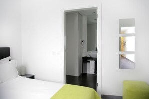 Quarto de hotel com cama, mesa de cabeceira, parede verde decorativa, banheiro com pia e espelho, e um pequeno espaço para sentar.