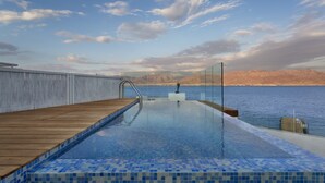 Piscina com deck de madeira e barreira de vidro, com vista para um corpo de água e montanhas ao longe.
