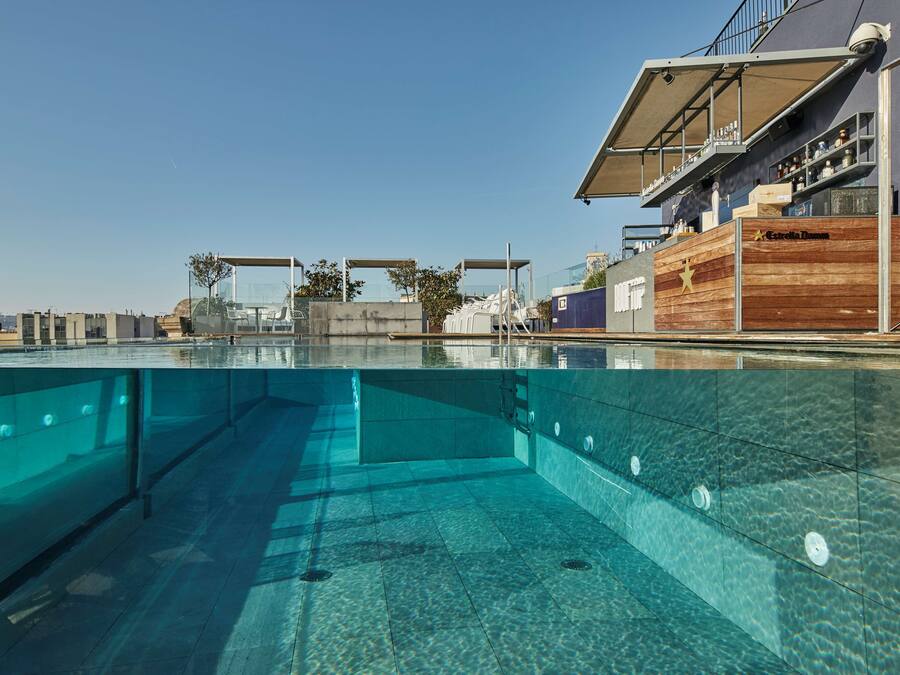Uma piscina translúcida com design moderno, rodeada por um deck de concreto e uma estrutura de madeira com o logotipo de uma estrela.