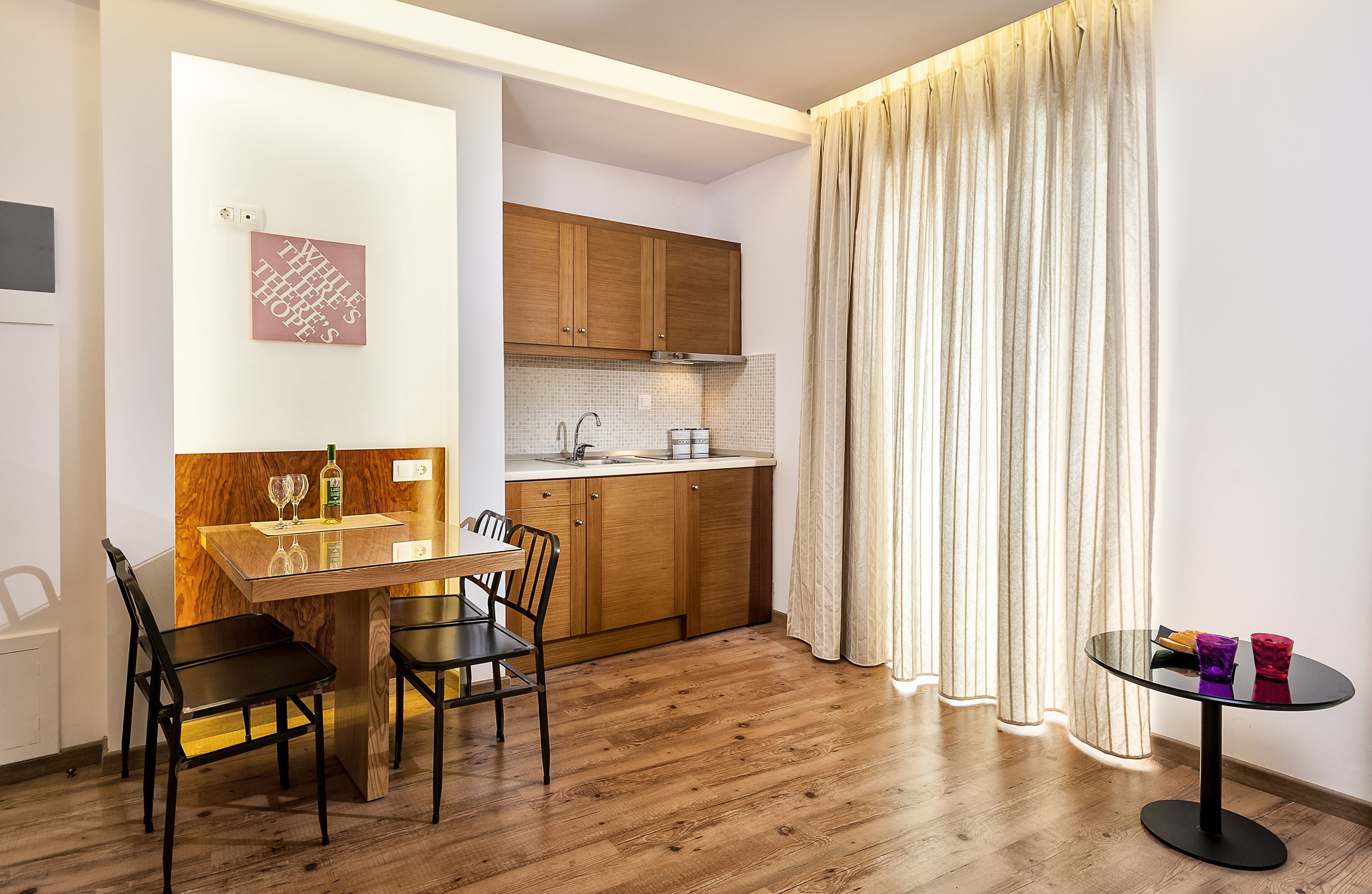 Cozinha compacta com armários de madeira, área de refeições com mesa e cadeiras, e uma pequena mesa redonda com copos.