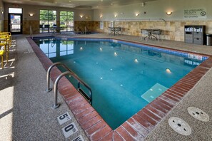 Piscina interior com borda em azulejo, posto de salva-vidas e área de estar com mesas e cadeiras.