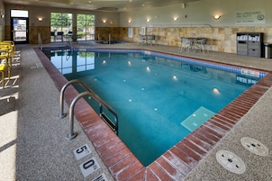 Piscina interior com borda em azulejo, posto de salva-vidas e área de estar com mesas e cadeiras.