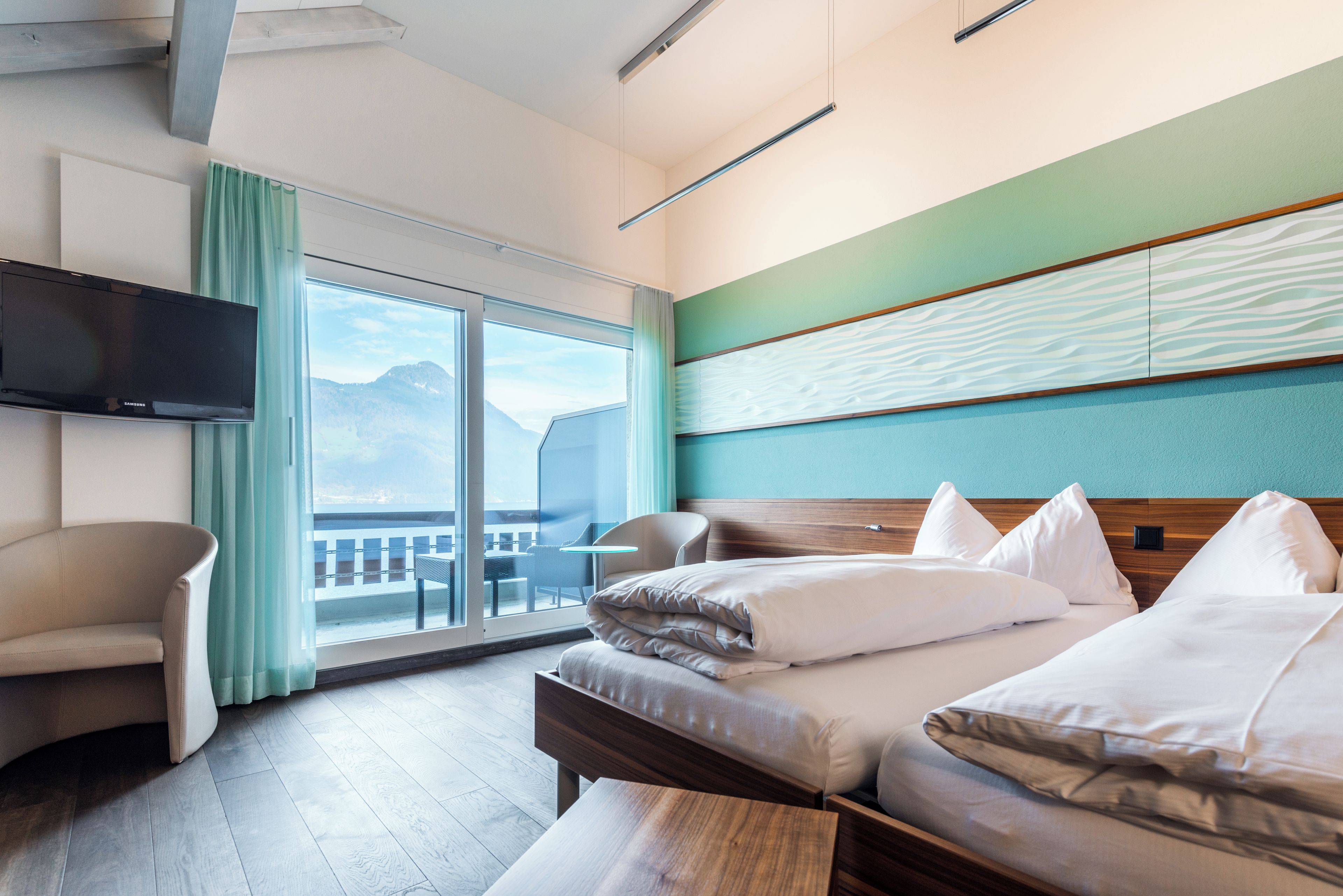 Quarto de hotel com cama, uma cadeira, televisão e vista para as montanhas pela janela.