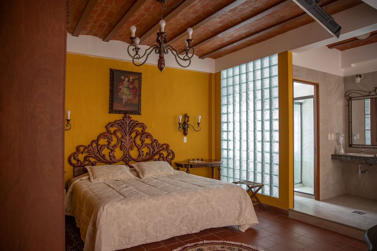 Quarto com cama grande, lustre, pintura na parede e banheiro com chuveiro e pia.