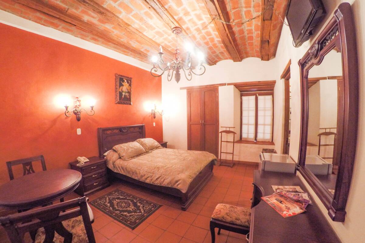 Um quarto com cama, escrivaninha, cadeira, cômoda, espelho e lustre.