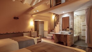 Quarto de hotel com duas camas, área de estar, banheiro com pia e espelho, e escada que leva a um andar superior.