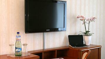 Quarto com televisão de tela plana, um laptop sobre a mesa, uma garrafa de água e uma orquídea em vaso.