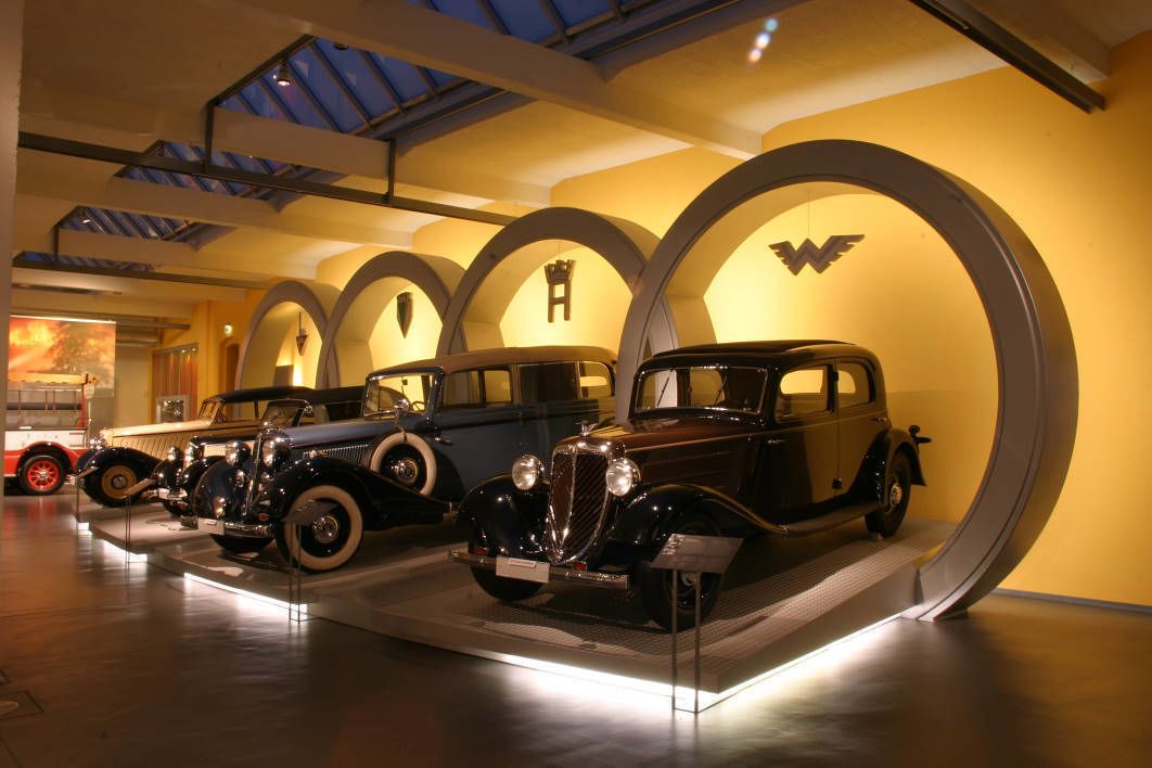 Exposição de museu com carros antigos exibidos em ambiente moderno, com tetos em arco e claraboias.