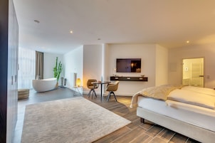 Quarto de hotel moderno com uma cama grande, área de estar com cadeiras, televisão de tela plana na parede e uma janela ampla com cortinas.