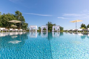 Piscina azul límpida com espreguiçadeiras e guarda-sóis brancos, cercada por árvores.
