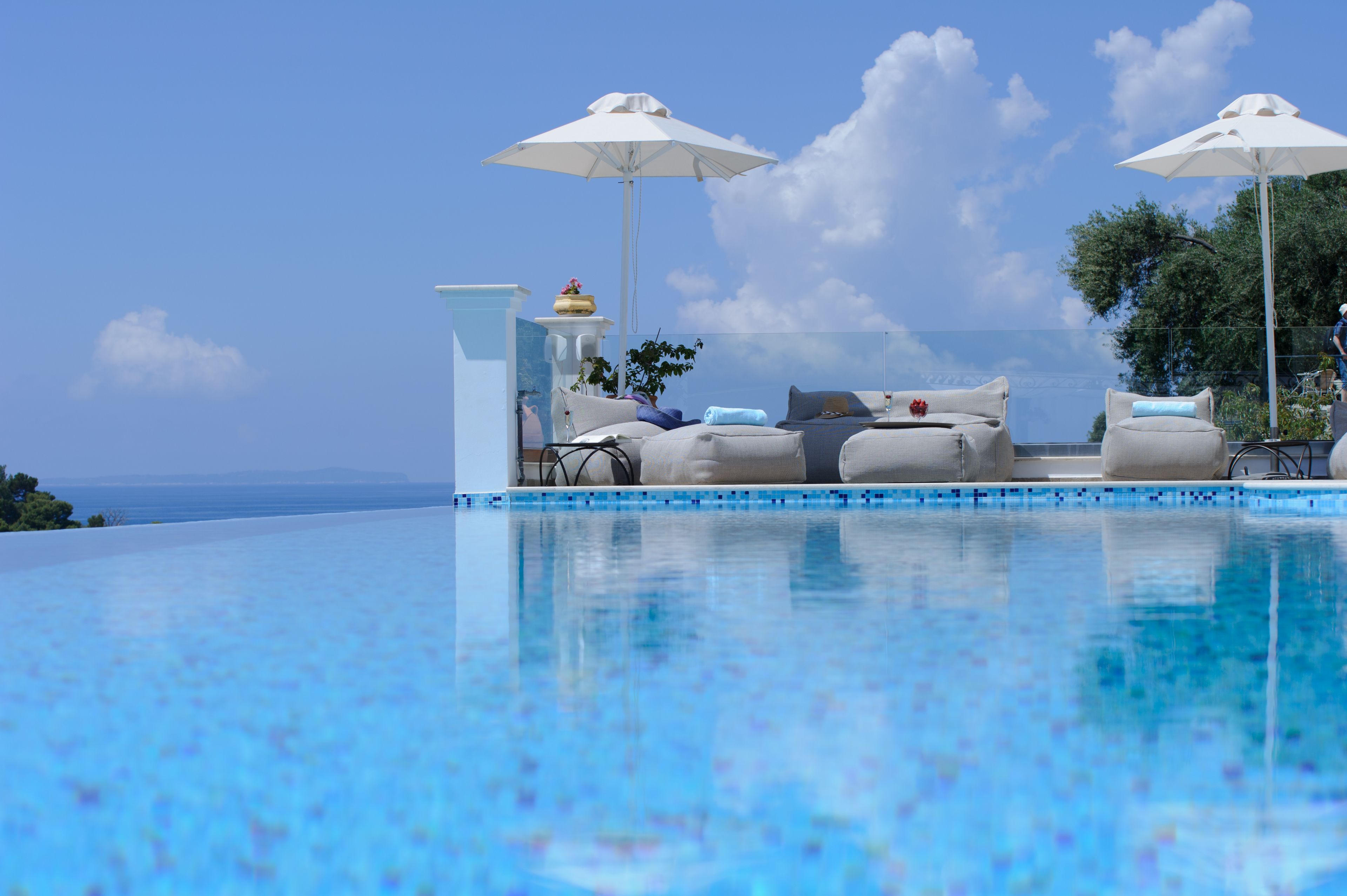 Piscina azul e límpida com espreguiçadeiras e guarda-sóis, uma fonte decorativa e vista para o mar.