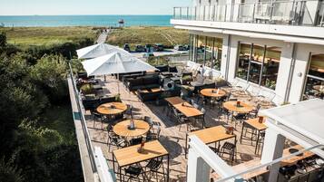 Um bar no terraço com mesas ao ar livre, vista para o mar e um edifício com grandes janelas.