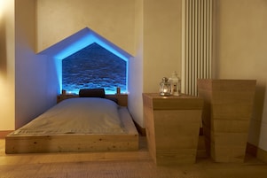 Quarto com teto inclinado, estrutura de cama de madeira, mesa de cabeceira e pequena mesa com um candeeiro.