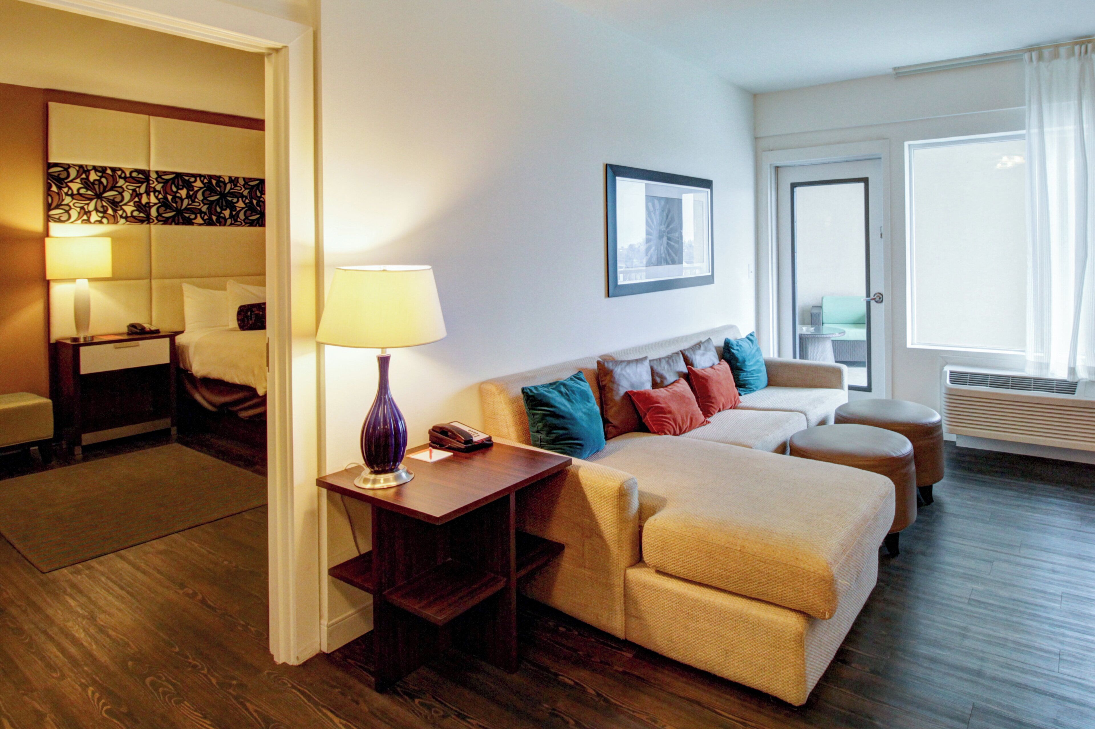 Quarto de hotel com sofá, mesa de cabeceira com luminária, cadeira, cama e janela com cortinas.