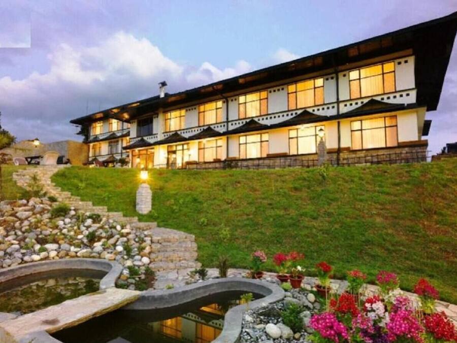 Um hotel moderno com uma estrutura grande e de vários andares, um jardim ajardinado com lago e um caminho de pedra que leva à entrada.