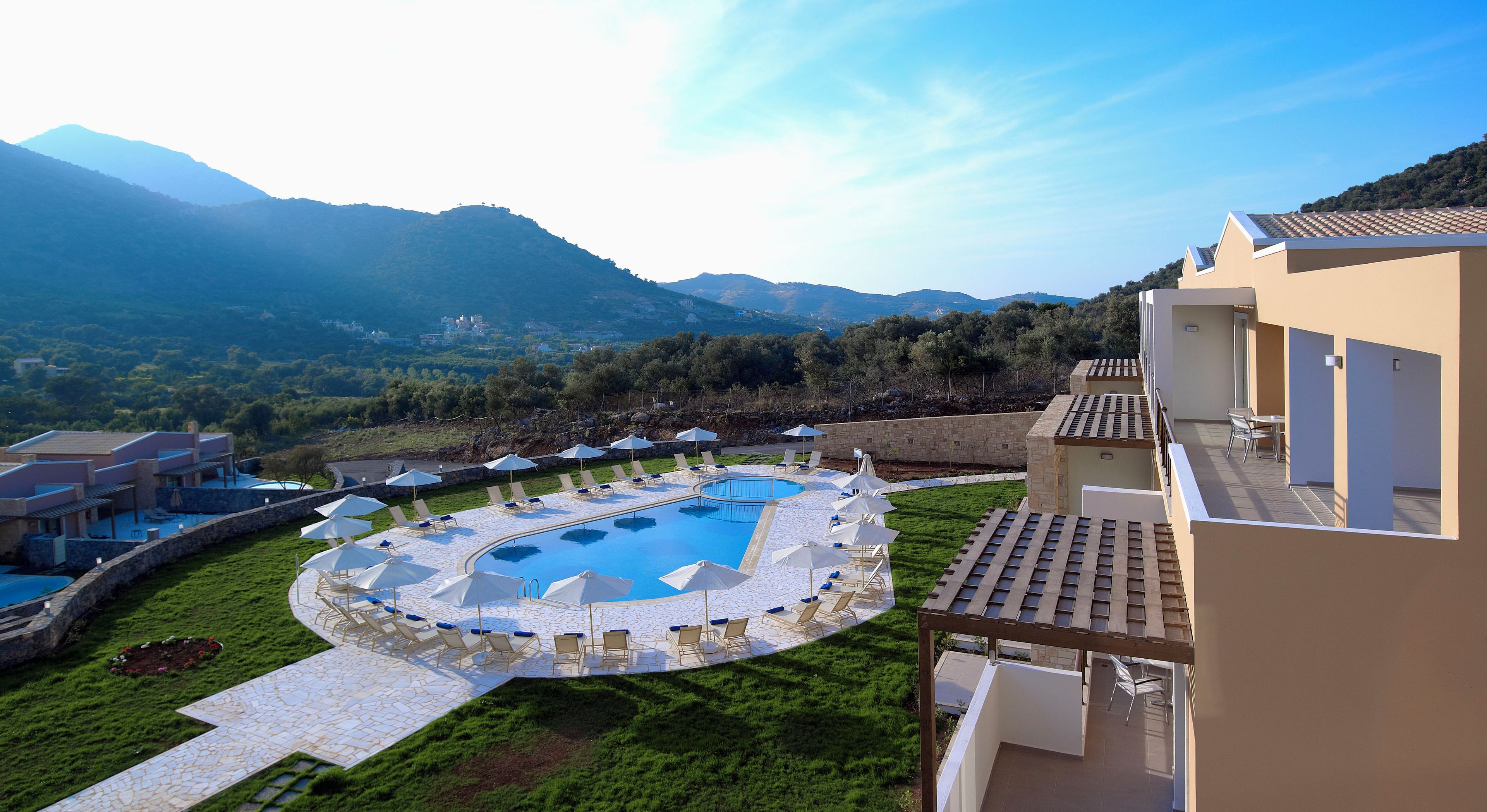 Área da piscina com espreguiçadeiras e guarda-sóis, cercada por um jardim paisagístico e vista para montanhas.