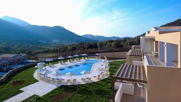 Área da piscina com espreguiçadeiras e guarda-sóis, cercada por um jardim paisagístico e vista para montanhas.