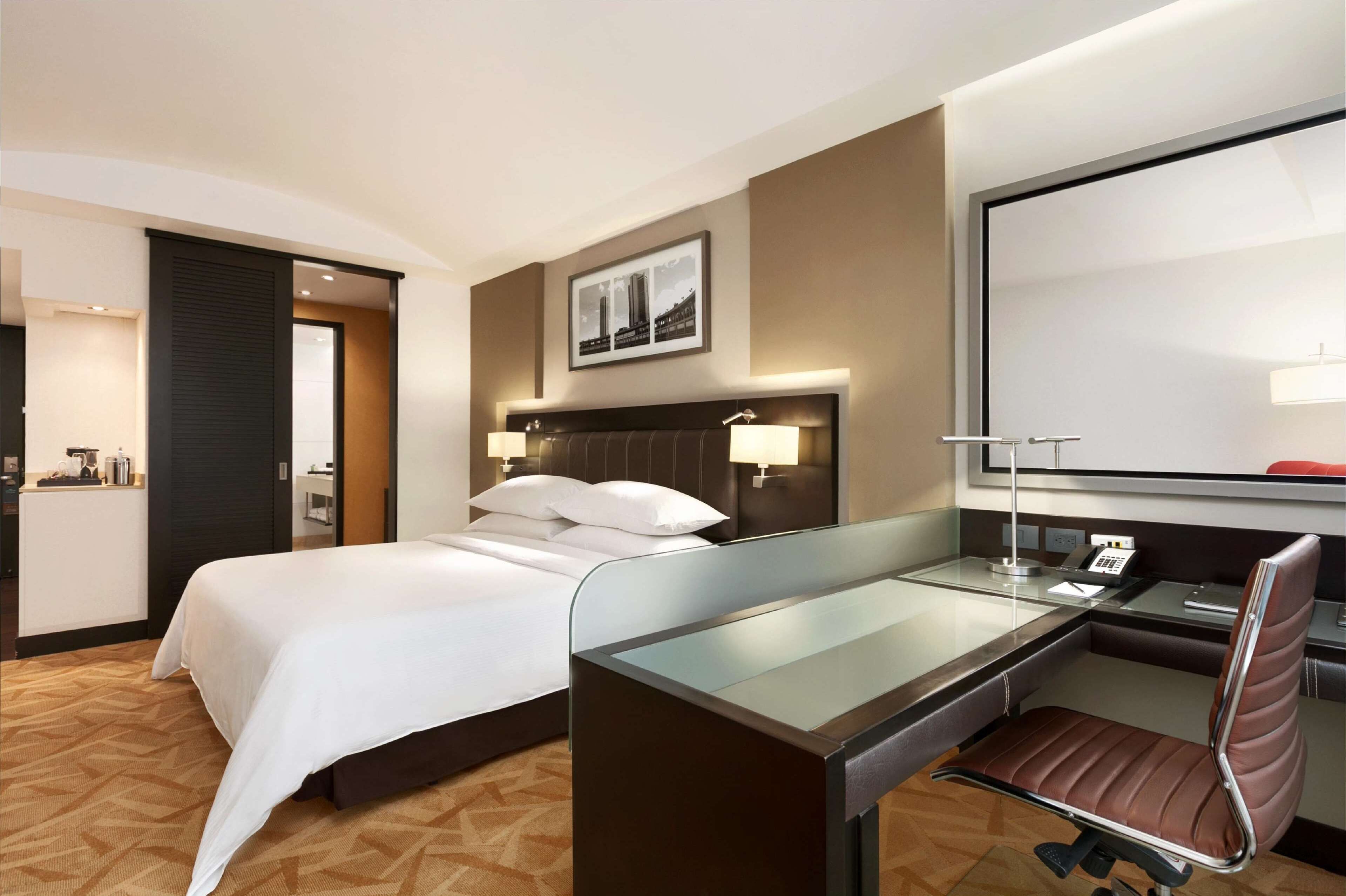 Quarto de hotel com cama, escrivaninha com cadeira, espelho grande e quadro na parede.