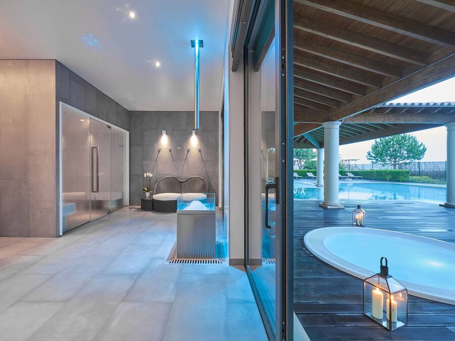 Uma área de spa moderna com sauna envidraçada, espaço de relaxamento com mesa de massagem e vista para uma piscina e área externa.