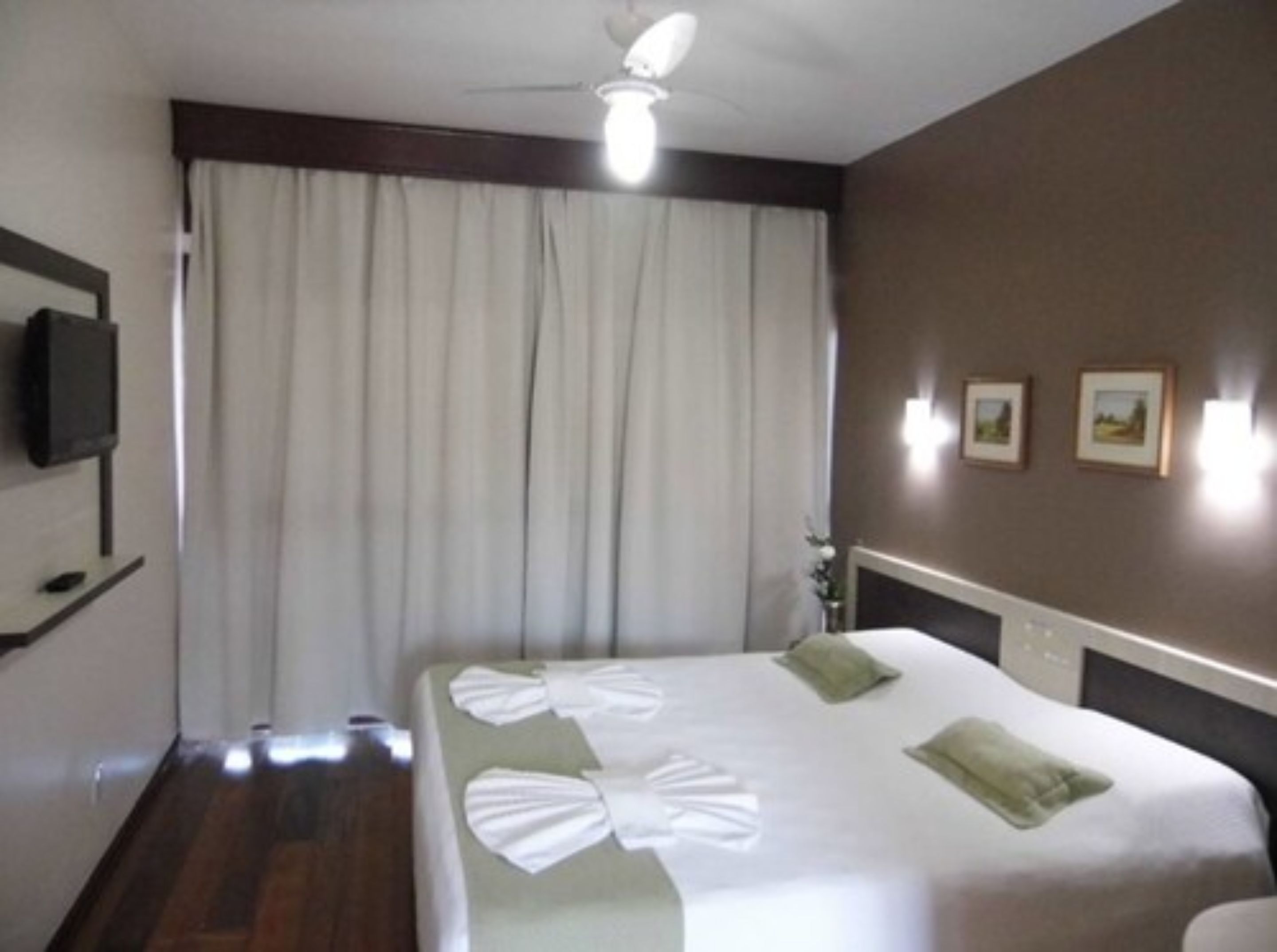 Quarto de hotel com uma cama, uma televisão de tela plana na parede e um ventilador de teto.