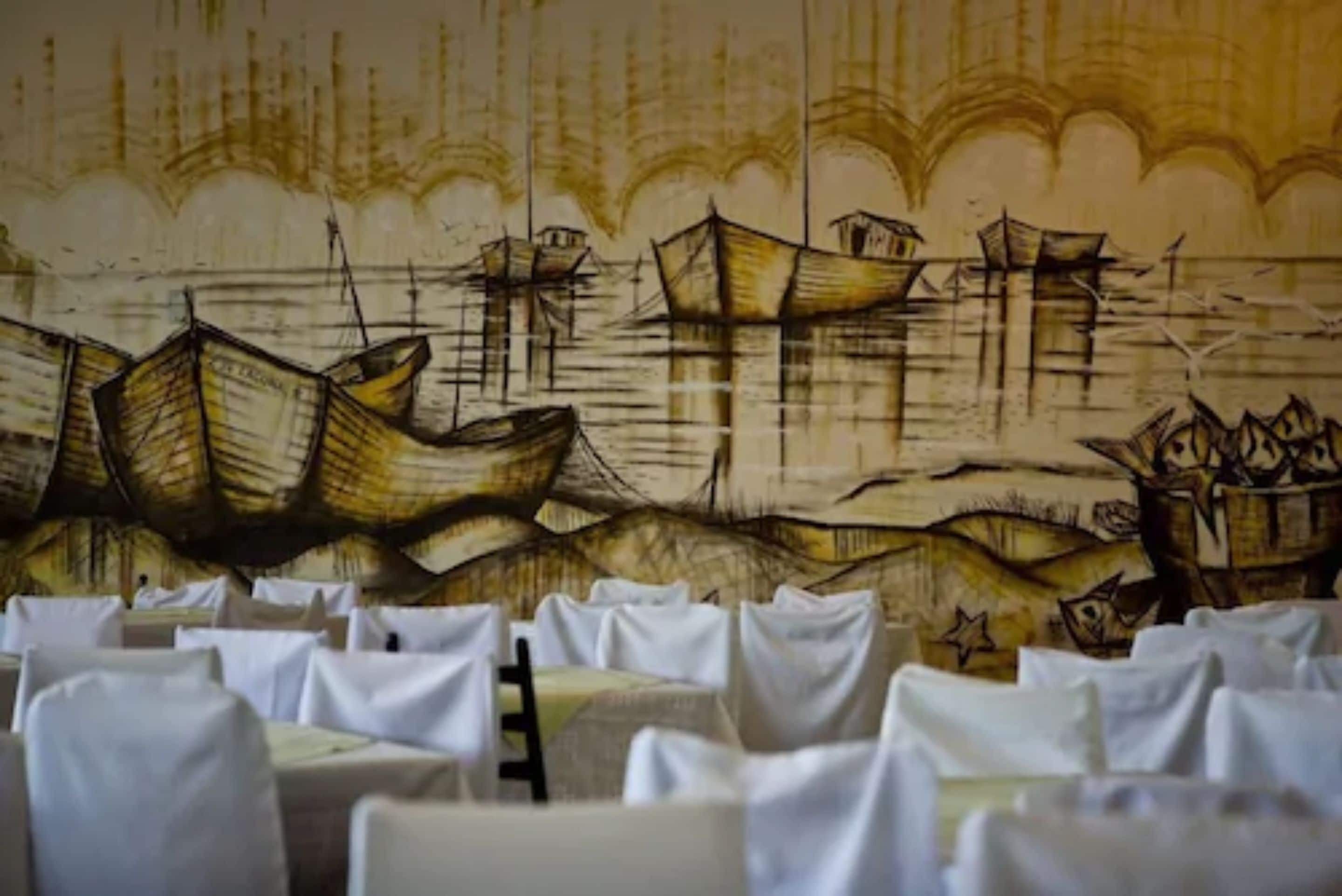Restaurante com mesas cobertas por toalhas brancas, um mural de barcos na água e um teto alto com detalhes em arco.