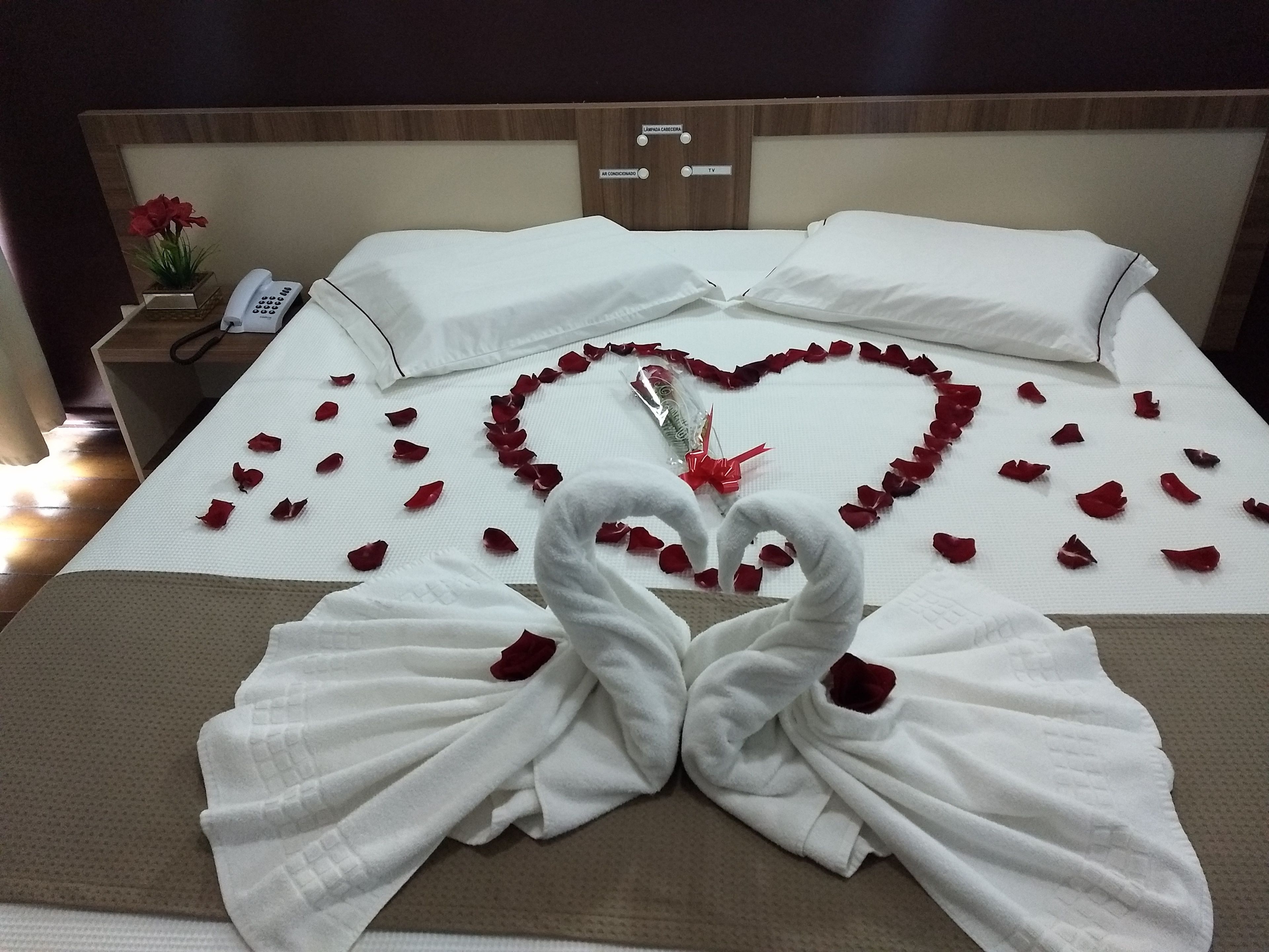 Quarto de hotel com uma cama bem arrumada, um arranjo de pétalas de rosa em formato de coração e duas toalhas em forma de cisne.