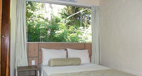 Quarto com cabeceira de madeira, cama com roupa de cama branca e vista para o verde pela janela.