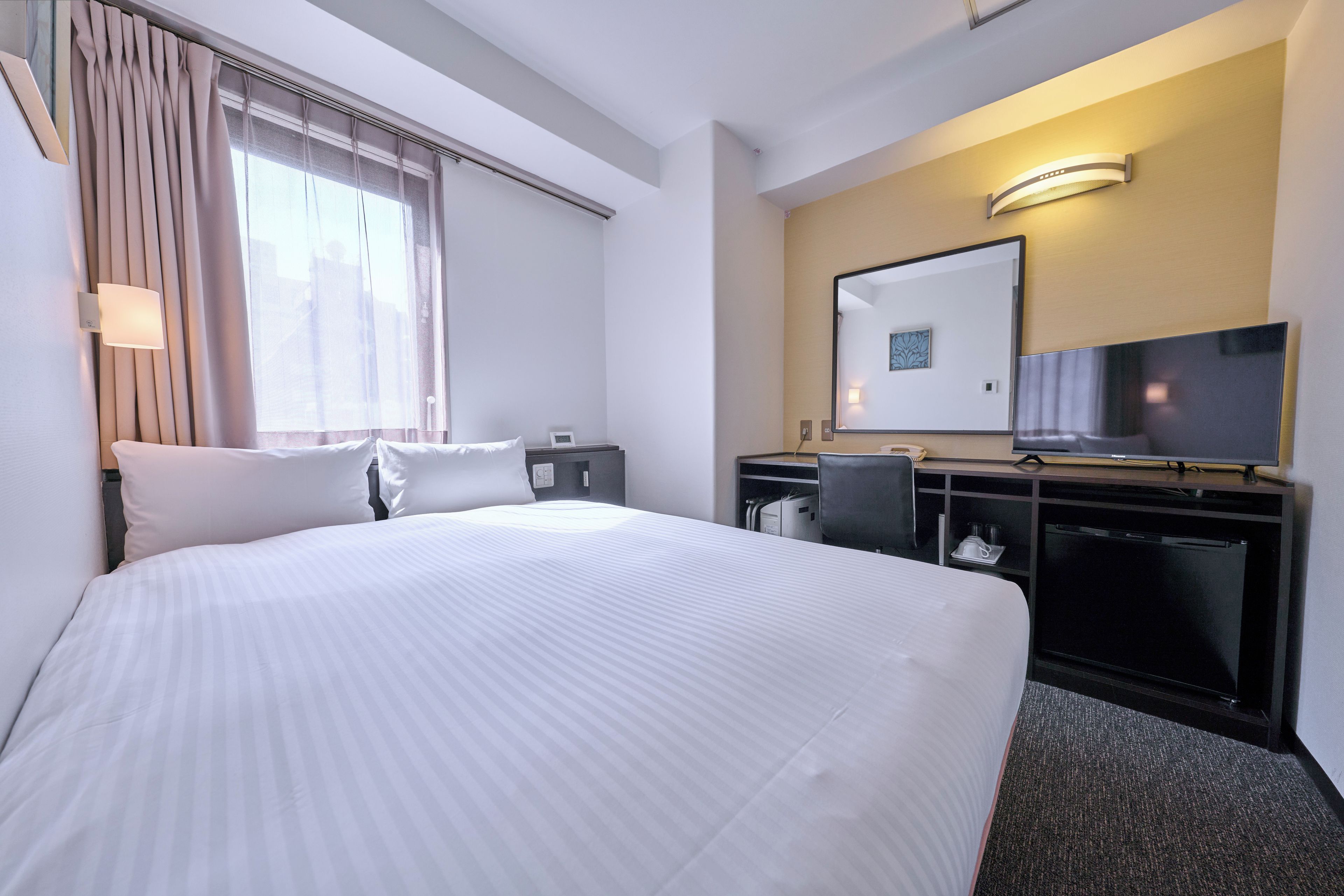 Quarto de hotel com uma cama grande, uma escrivaninha com cadeira, televisão e espelho.