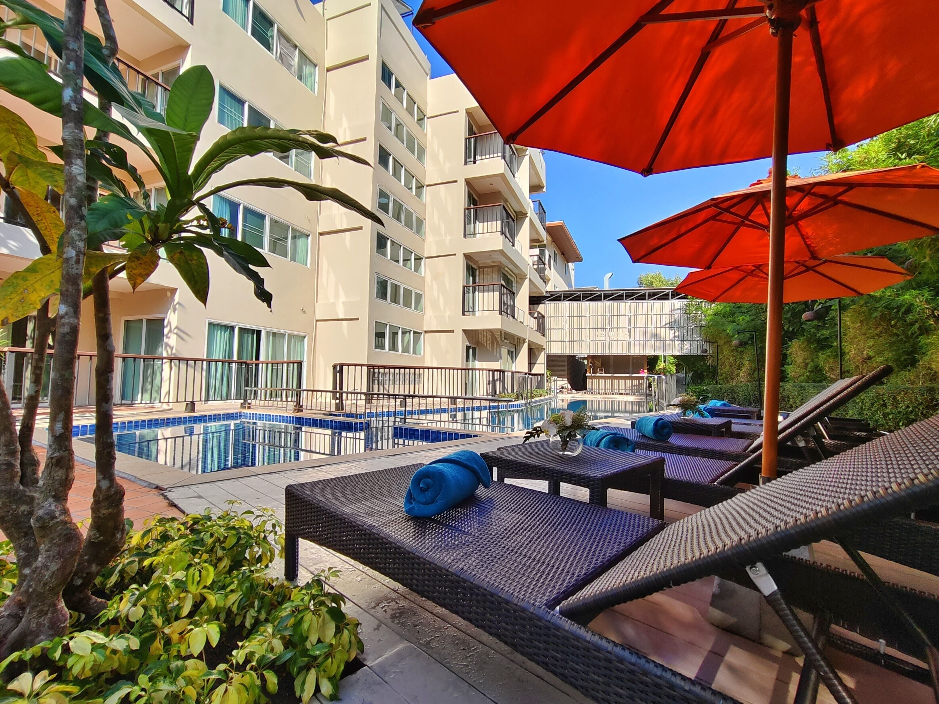 Uma área à beira da piscina com espreguiçadeiras e guarda-sóis, cercada por um edifício com várias varandas.