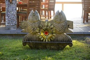 Escultura de pedra em formato de peixe com um arranjo floral central, colocada sobre grama em uma área de refeições ao ar livre com cadeiras e mesas de madeira.