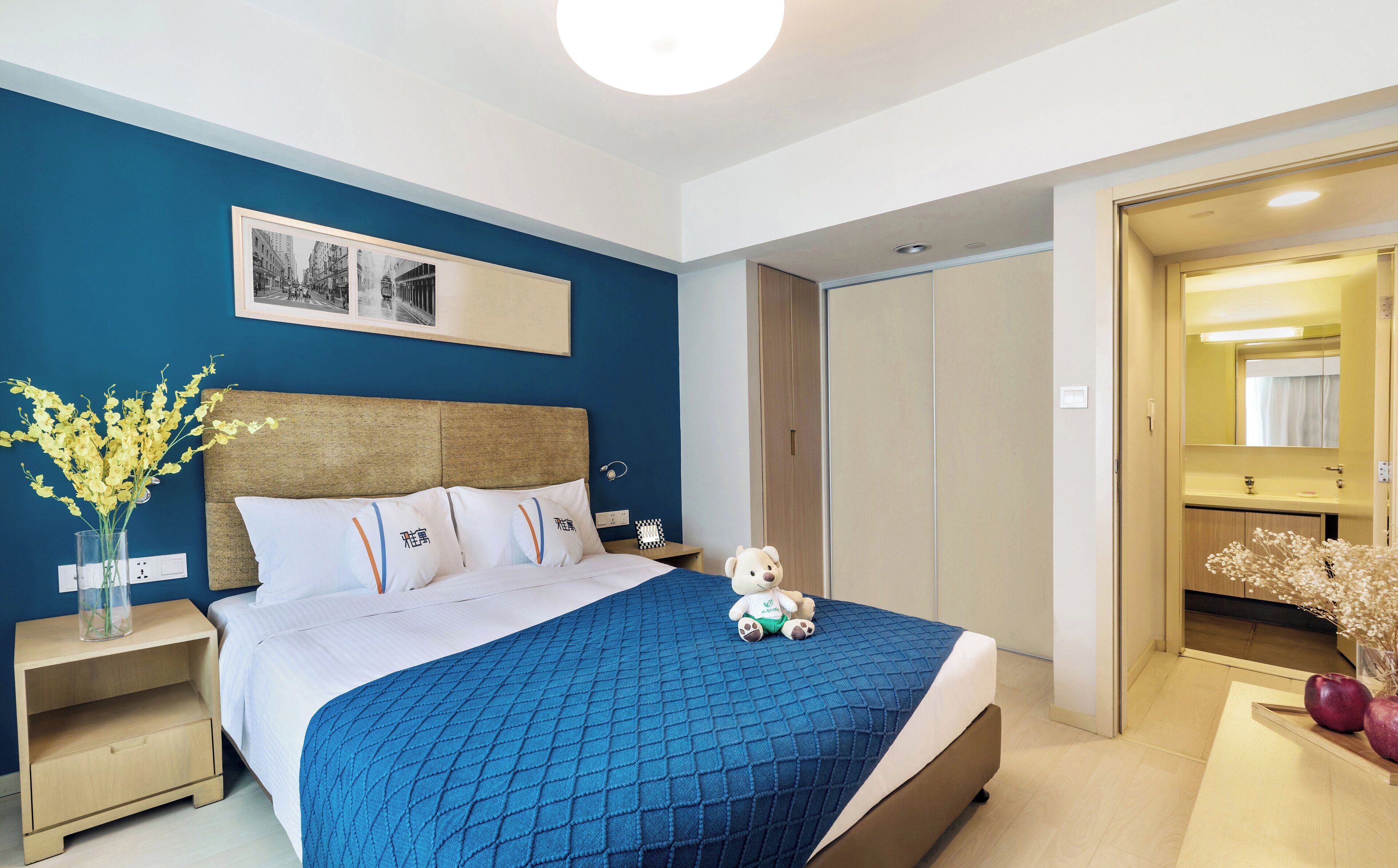 Quarto de hotel bem arrumado com cama, mesa de cabeceira com um vaso de flores, um quadro na parede e um ursinho na cama.