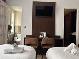 Quarto de hotel com duas camas, uma secretária com cadeiras, uma televisão presa à parede e um banheiro visível por uma porta aberta.