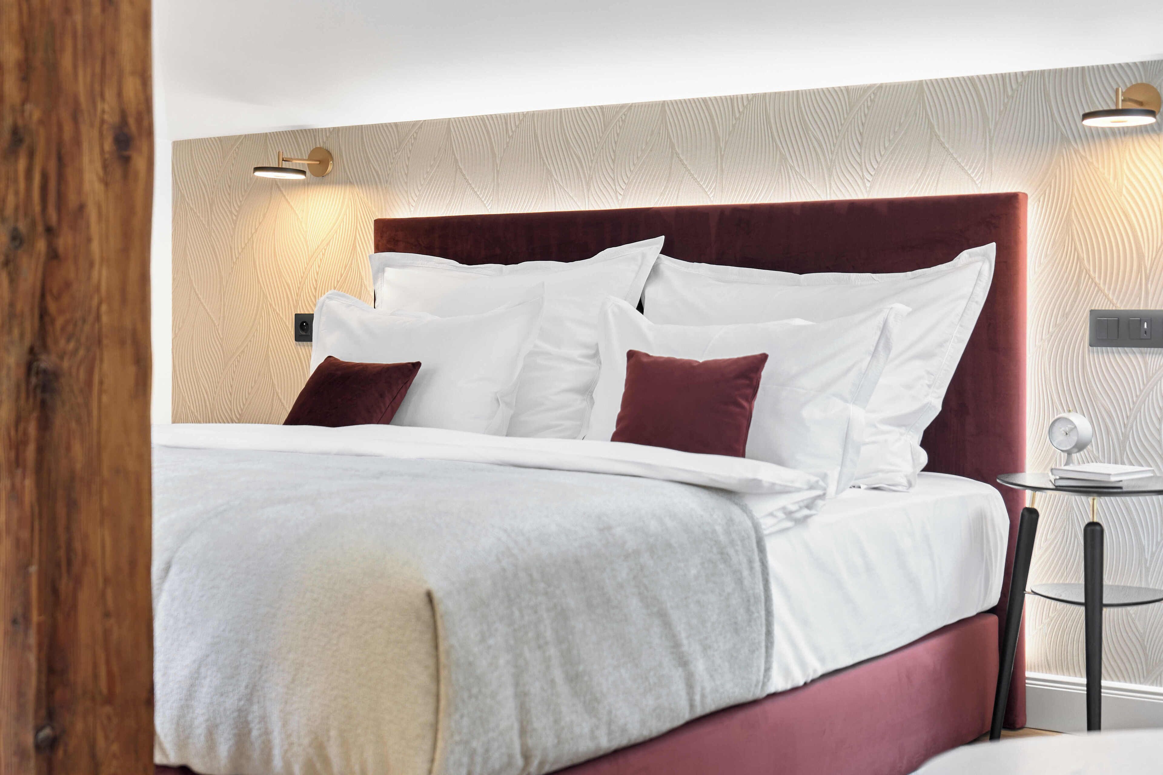 Cama com lençóis brancos e detalhes vermelhos, cabeceira de madeira e uma pequena mesa de cabeceira com abajur.