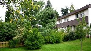 Zona residencial com gramado bem cuidado, diversos tipos de árvores e um edifício com telhado plano e várias janelas.