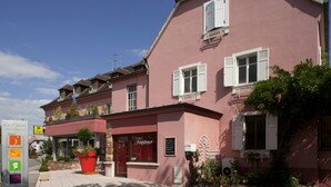 Um edifício cor-de-rosa com o letreiro 'Traiteur', um vaso de planta vermelho grande e uma placa de sinalização listando comodidades como hotel, restaurante e boutique.